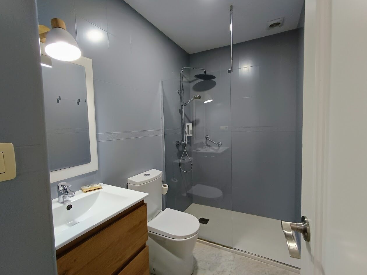 Um banheiro moderno com pia branca, espelho e vaso sanitário, chuveiro com porta de vidro e toucador de madeira.