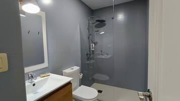 Um banheiro moderno com pia branca, espelho e vaso sanitário, chuveiro com porta de vidro e toucador de madeira.