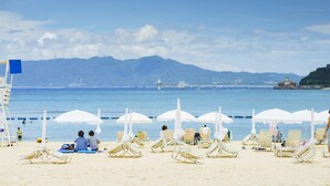 Uma praia com fileiras de espreguiçadeiras e guarda-sóis, uma torre salva-vidas e vista para o mar e montanhas ao longe.