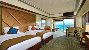 Quarto de hotel com duas camas, varanda com vista para o mar e área de estar com mesa e cadeiras.