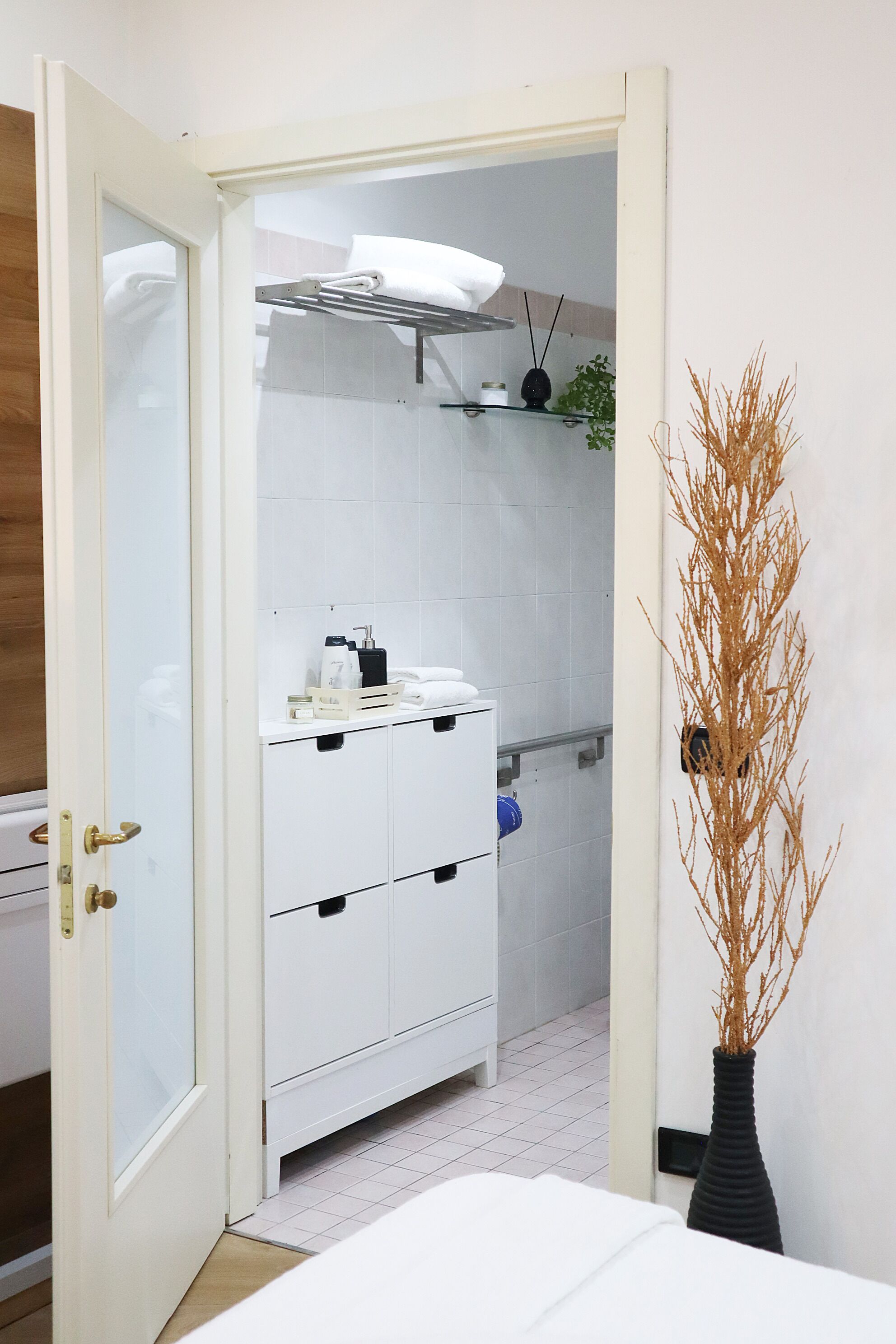 Casa de banho com lavatório branco, porta-toalhas e um vaso preto com plantas secas.