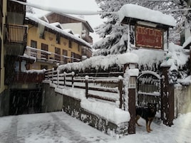 Um portão de entrada coberto de neve com cerca de madeira, uma placa com a inscrição "LES LACS Residence" e um cão próximo.