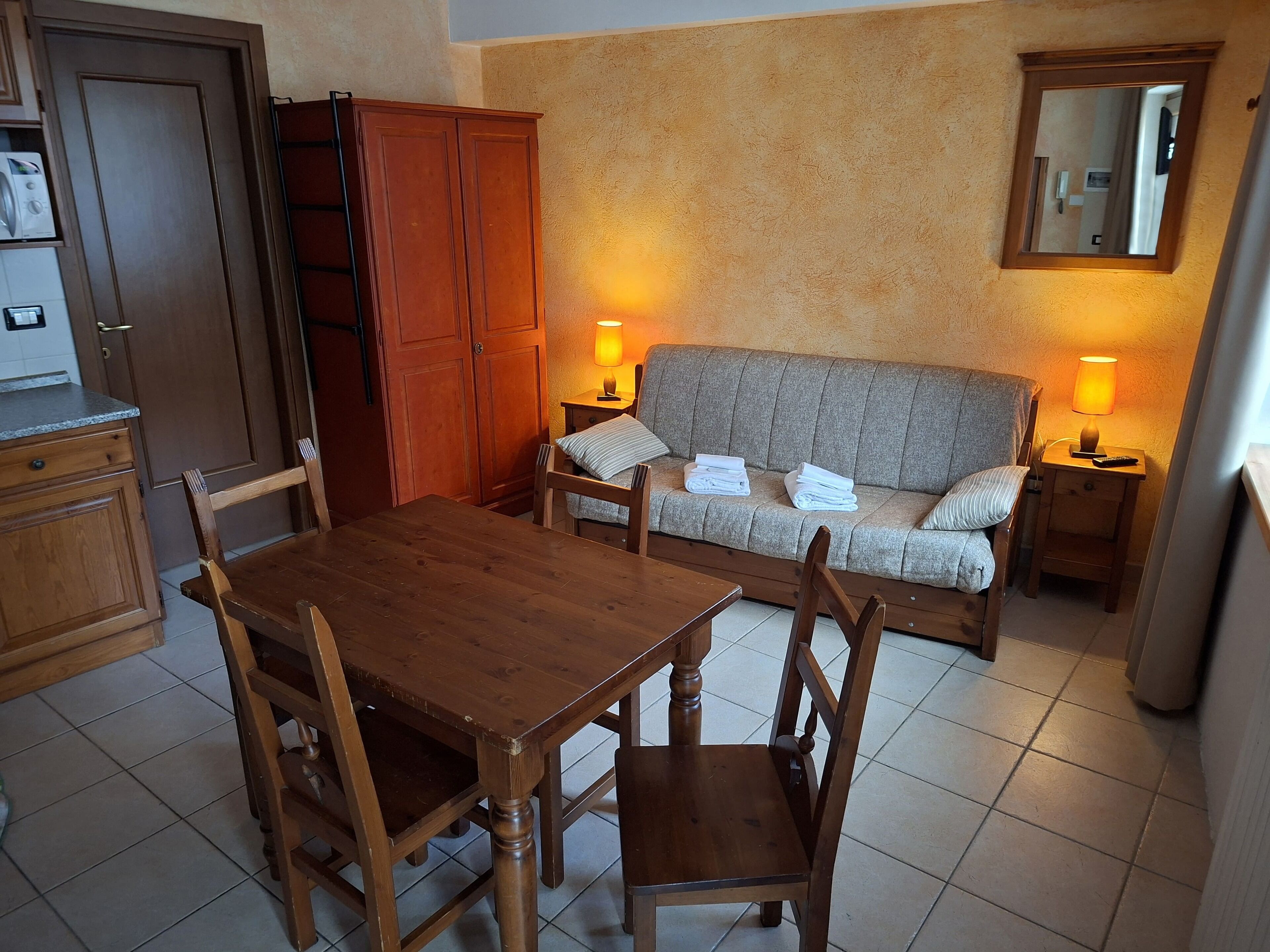 Uma sala de estar aconchegante com uma mesa de jantar de madeira e cadeiras, um sofá cinza com almofadas e duas luminárias de mesa.