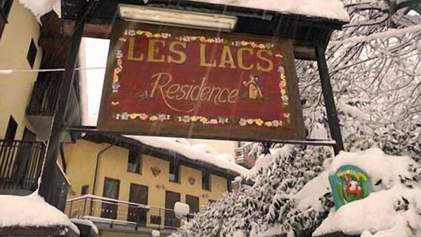Placa de 'LES LACS Residence', com telhado coberto de neve e um edifício ao fundo.