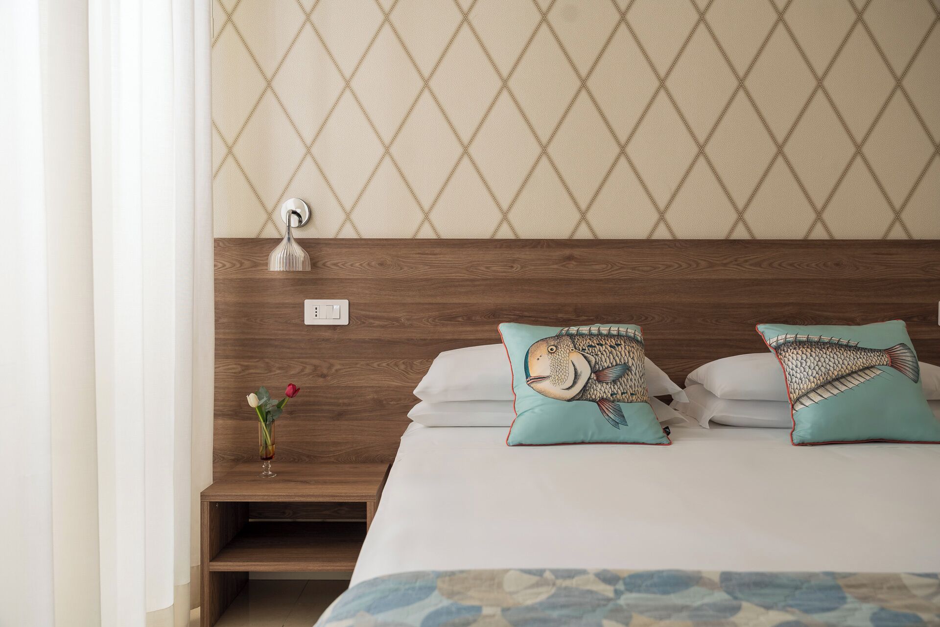 Quarto de hotel com cabeceira de madeira, mesa de cabeceira com um vaso de flores e duas almofadas decorativas com desenhos de peixes.
