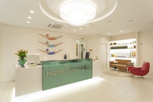 Uma área de recepção de hotel com design moderno, apresentando um balcão de recepção, um candelabro, uma poltrona vermelha e uma estante com livros e objetos decorativos.