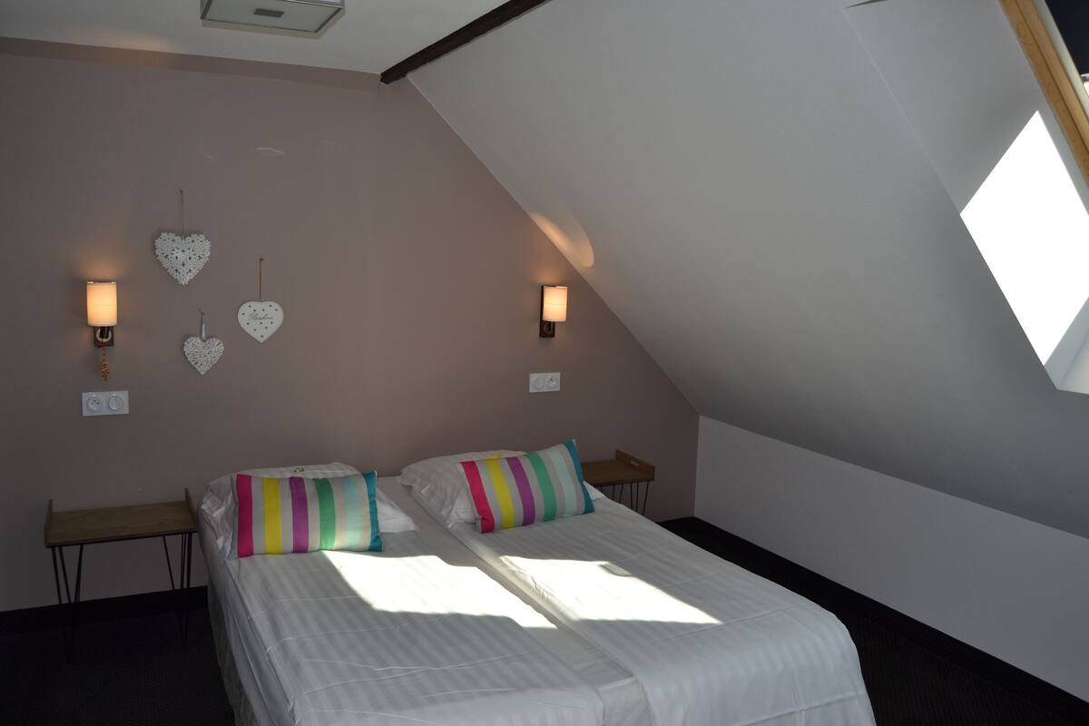 Quarto com teto inclinado, duas mesas de cabeceira, cama com almofadas listradas, luminárias fixadas na parede e decorações em formato de coração.