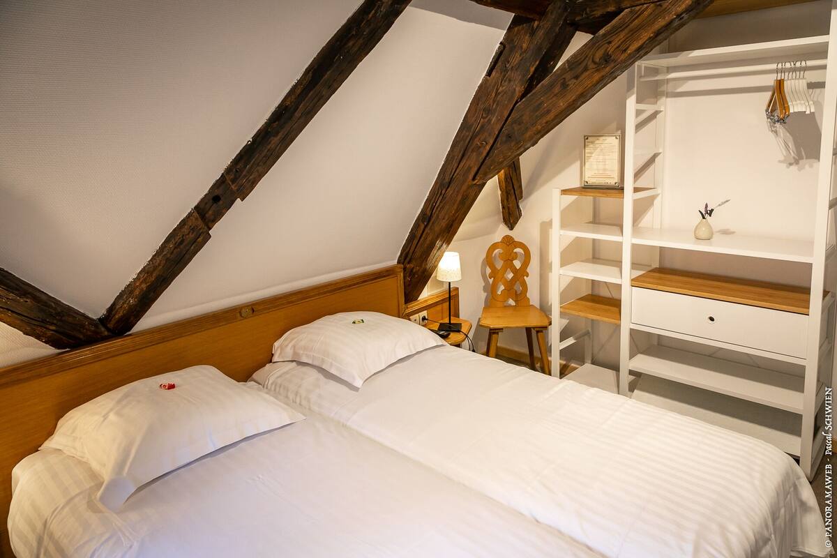 Quarto com teto inclinado, vigas de madeira, cama com roupa de cama branca, mesa de cabeceira com luminária e estante branca com um vaso.