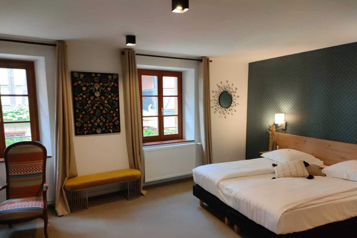 Quarto com cama, um banquinho, uma cadeira, janela e luminária fixada na parede.