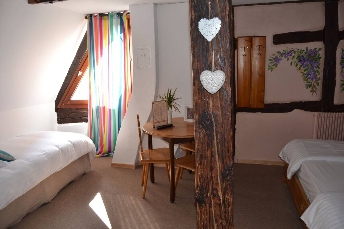 Quarto com duas camas, uma mesa de madeira, uma cadeira e uma janela com cortina colorida.