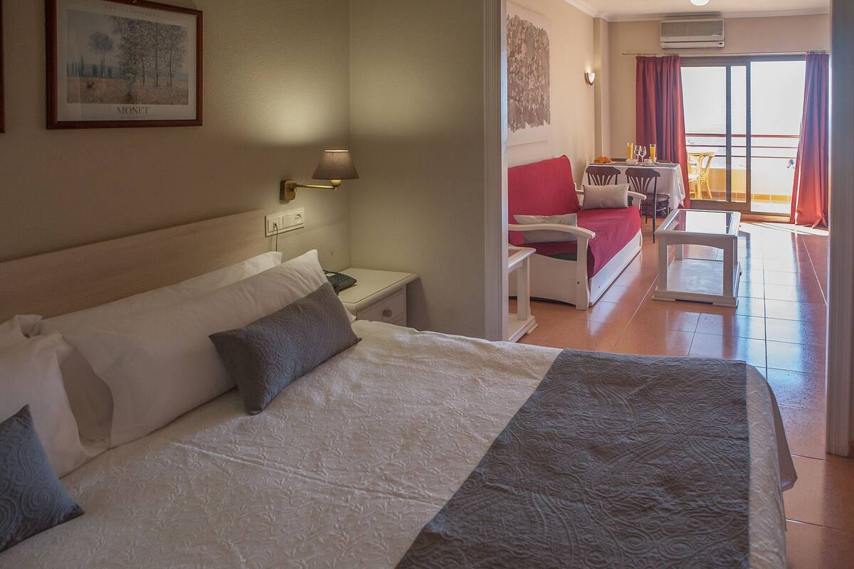 Quarto de hotel com uma cama grande, uma escrivaninha, uma cadeira e varanda com cortinas vermelhas.