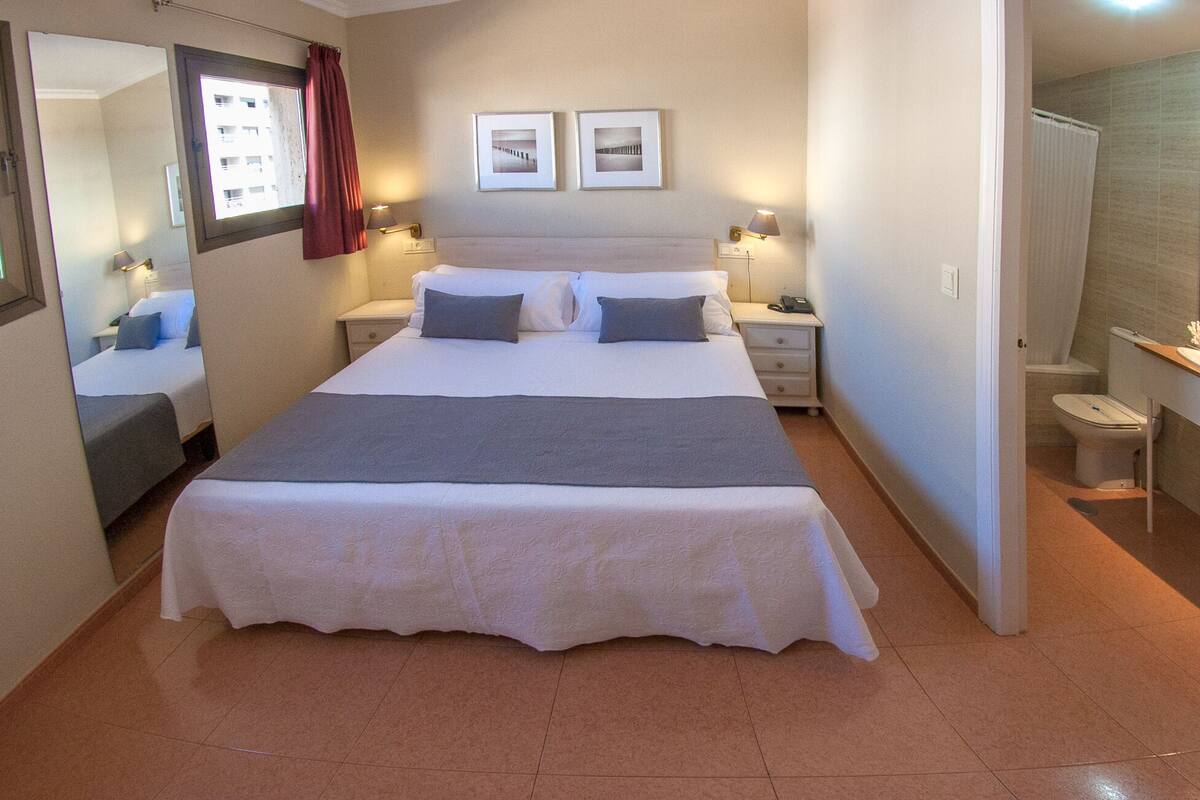Quarto de hotel com uma cama grande, duas mesas de cabeceira, um criado-mudo, um espelho e um banheiro visível por uma porta aberta.