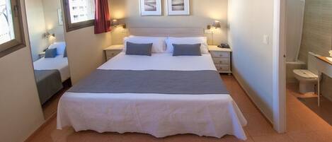 Quarto de hotel com uma cama grande, duas mesas de cabeceira, um criado-mudo, banheiro com vaso sanitário e pia, e duas imagens emolduradas na parede.