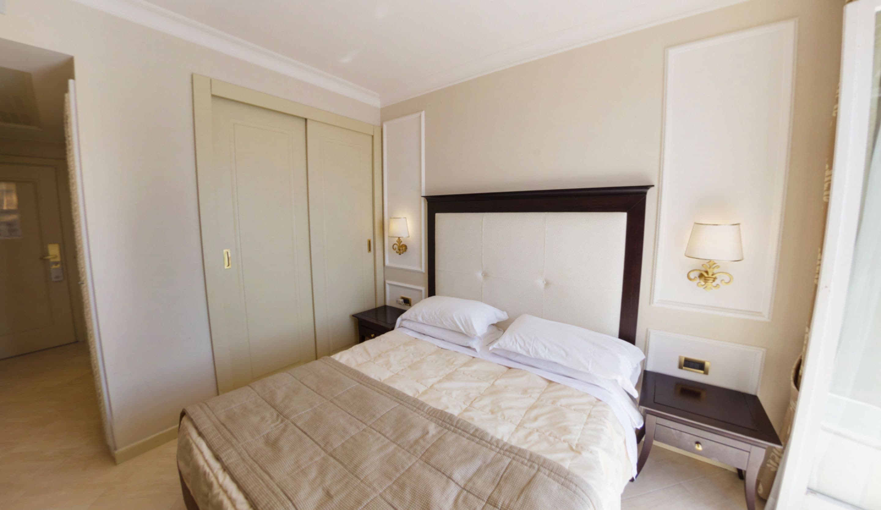 Quarto de hotel com uma cama grande, um guarda-roupa, uma mesa de cabeceira com abajur e uma luminária na parede.