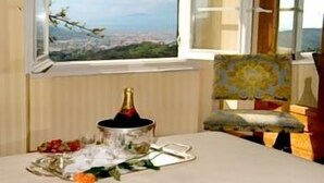 Quarto com vista para a paisagem urbana através de uma janela, uma mesa com uma garrafa e flores, e uma cadeira com almofada estampada.