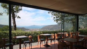 Área externa para refeições com mesas e cadeiras, grandes janelas com vista para a cidade e montanhas.