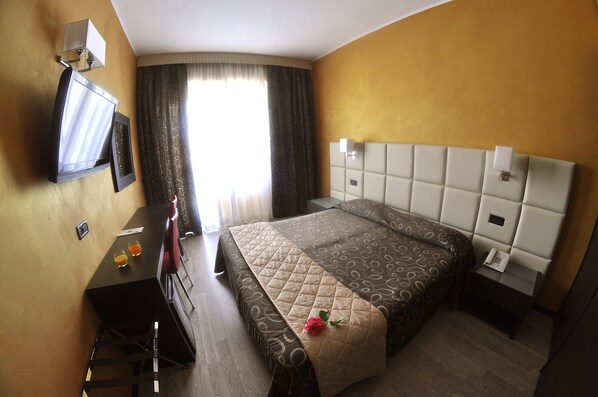 Quarto de hotel com cama, escrivaninha, cadeira, televisão e janela com cortinas.