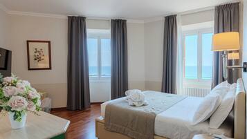 Quarto de hotel com cama, luminária, janela com cortinas, quadro na parede e vaso com flores na mesinha de cabeceira.