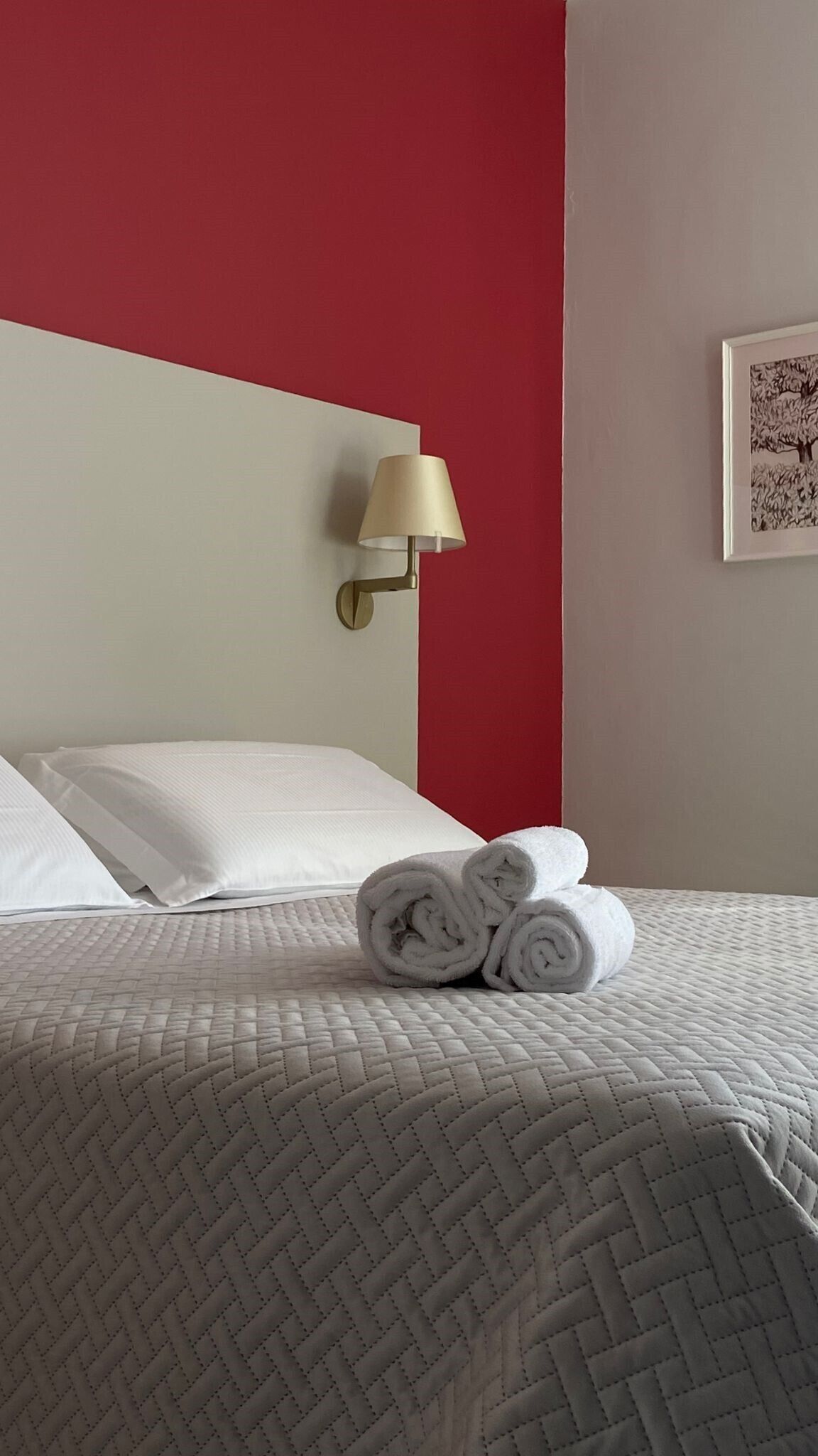 Uma cama bem arrumada com um edredom acolchoado, uma toalha enrolada e uma luminária na parede.