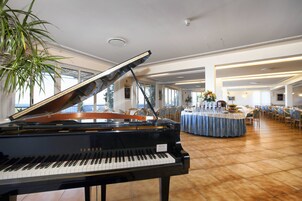 Um piano de cauda em sala ampla, com teto alto, janelas grandes e um espaço de jantar com mesas redondas.