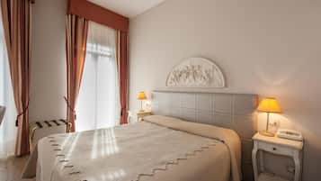 Quarto de hotel com cama, duas luminárias de cabeceira, mesa de cabeceira com telefone, uma cadeira e janela com cortinas.