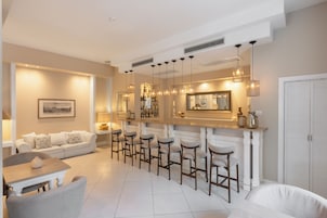 Uma sala de estar moderna com um sofá, uma mesa de jantar e banquetas altas. Há uma área de cozinha com um balcão e um espelho.