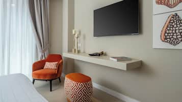 Quarto de hotel moderno com cama, televisão fixada na parede, um pequeno espaço de estar com poltrona e apoio para os pés, e uma estante com objetos decorativos.