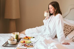 Uma pessoa vestindo um roupão branco sentada na cama com itens de café da manhã, incluindo frutas, panquecas e uma xícara.