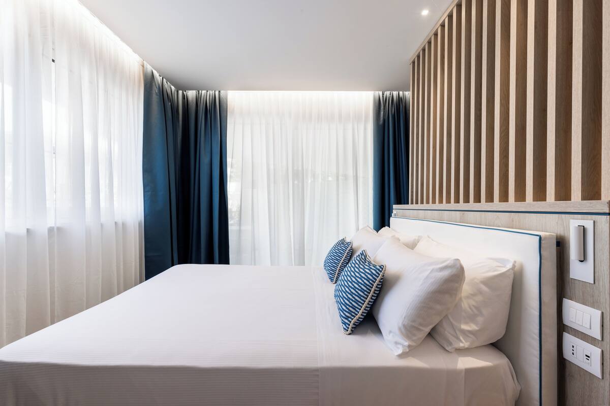 Quarto de hotel moderno com uma cama grande, roupa de cama branca e cabeceira de madeira.