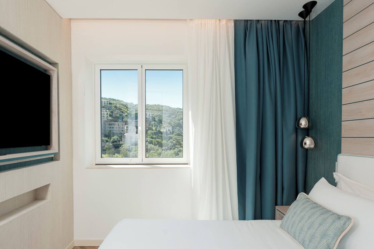 Quarto de hotel com cama, televisão, janela com cortinas e vista para edifícios.