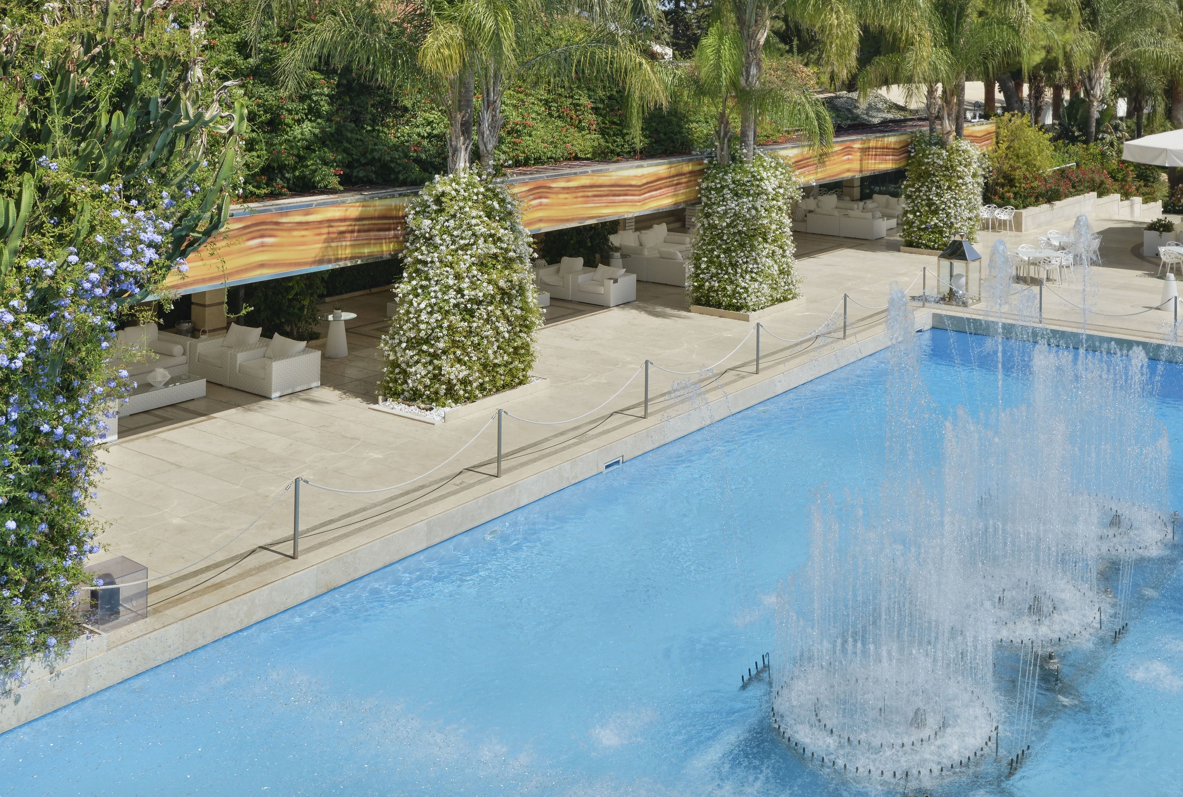 Uma piscina com uma fonte, rodeada por mobiliário de jardim branco e vegetação.