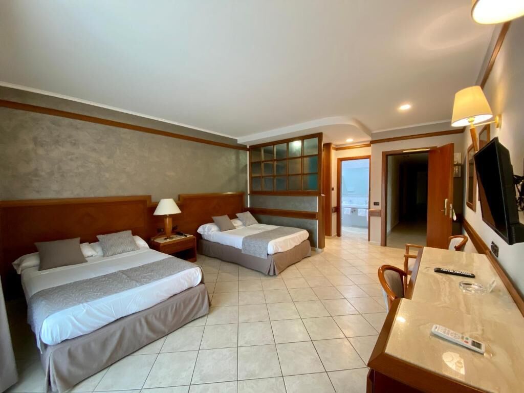 Quarto de hotel com duas camas, uma escrivaninha, televisão e banheiro visível por uma porta aberta.