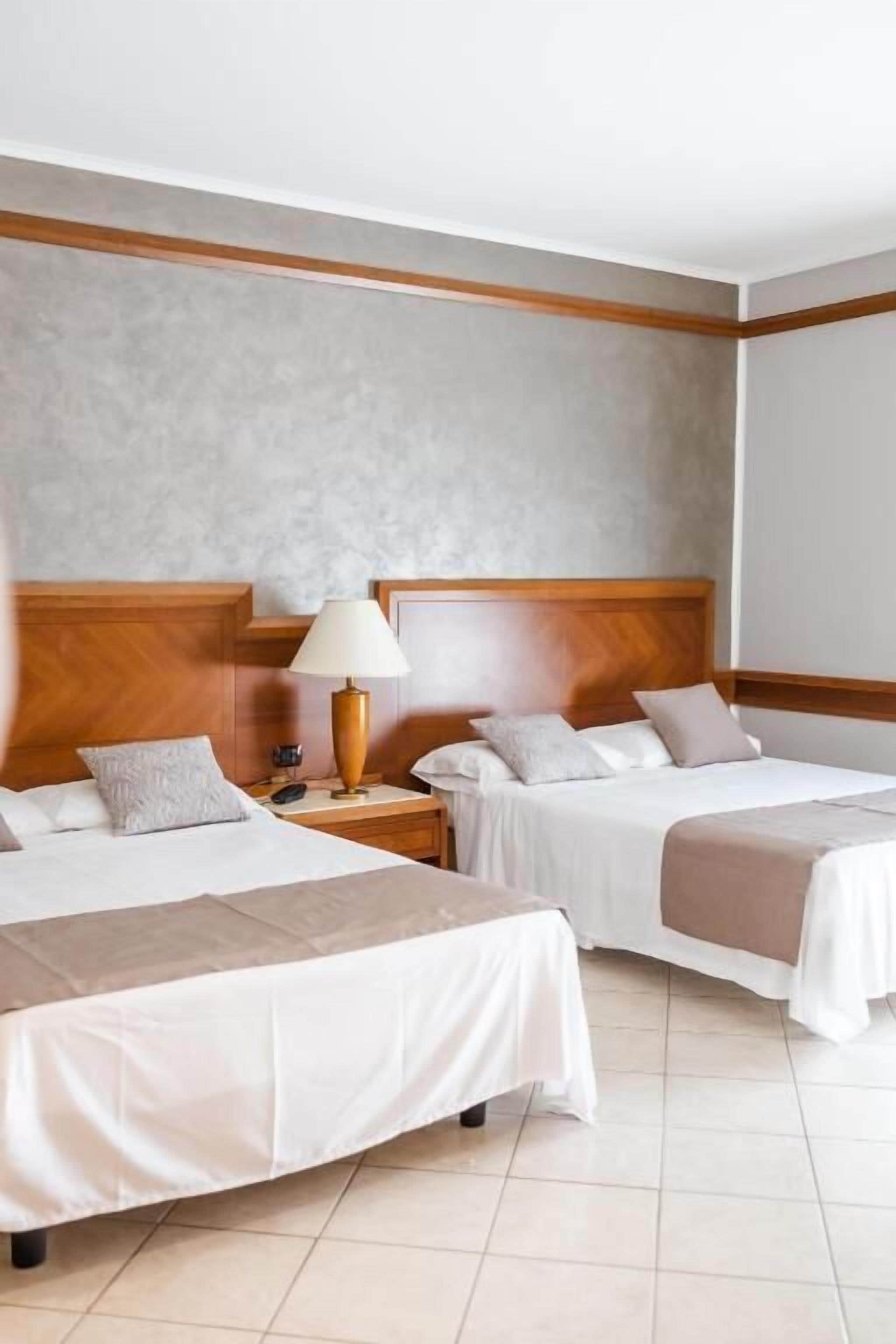 Quarto de hotel com duas camas, uma cabeceira de madeira, mesa de cabeceira com abajur e parede lisa.