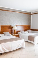 Quarto de hotel com duas camas, uma cabeceira de madeira, mesa de cabeceira com abajur e parede lisa.