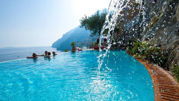 Uma piscina com águas azuis cristalinas, cascata e vista para o mar e montanhas.