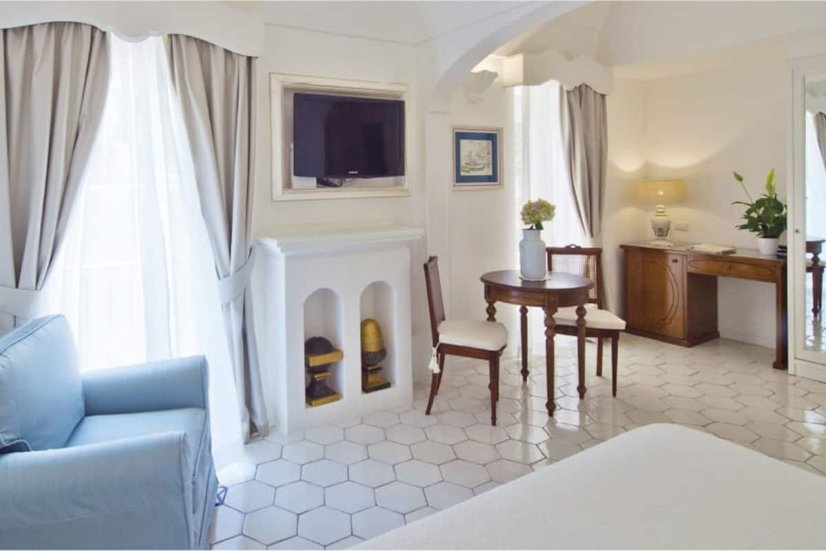 Quarto de hotel com uma cama, uma poltrona azul, uma escrivaninha de madeira e uma mesa de jantar.
