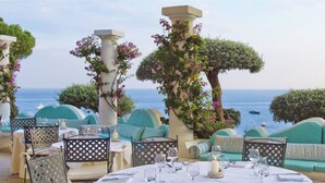 Uma área de refeições ao ar livre com mesas e cadeiras, vista para o mar e plantas decorativas.