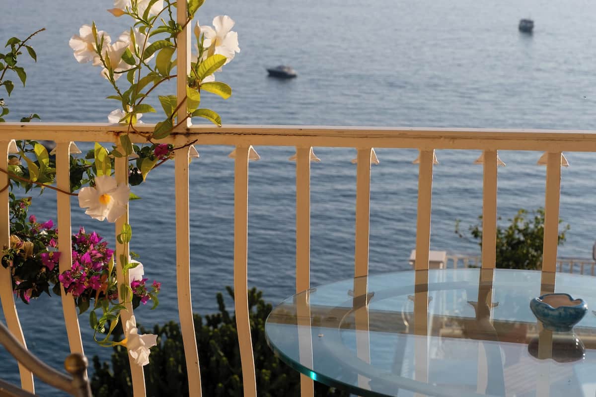 Varanda com mesa e cadeiras de vidro, com vista para um mar tranquilo com barcos.