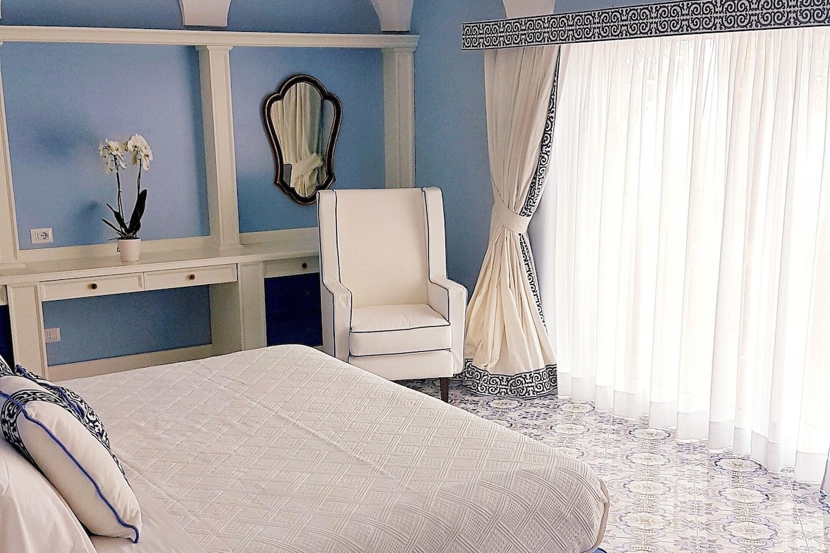 Quarto com cama grande, mesa de cabeceira branca, cadeira, cômoda e lustre.