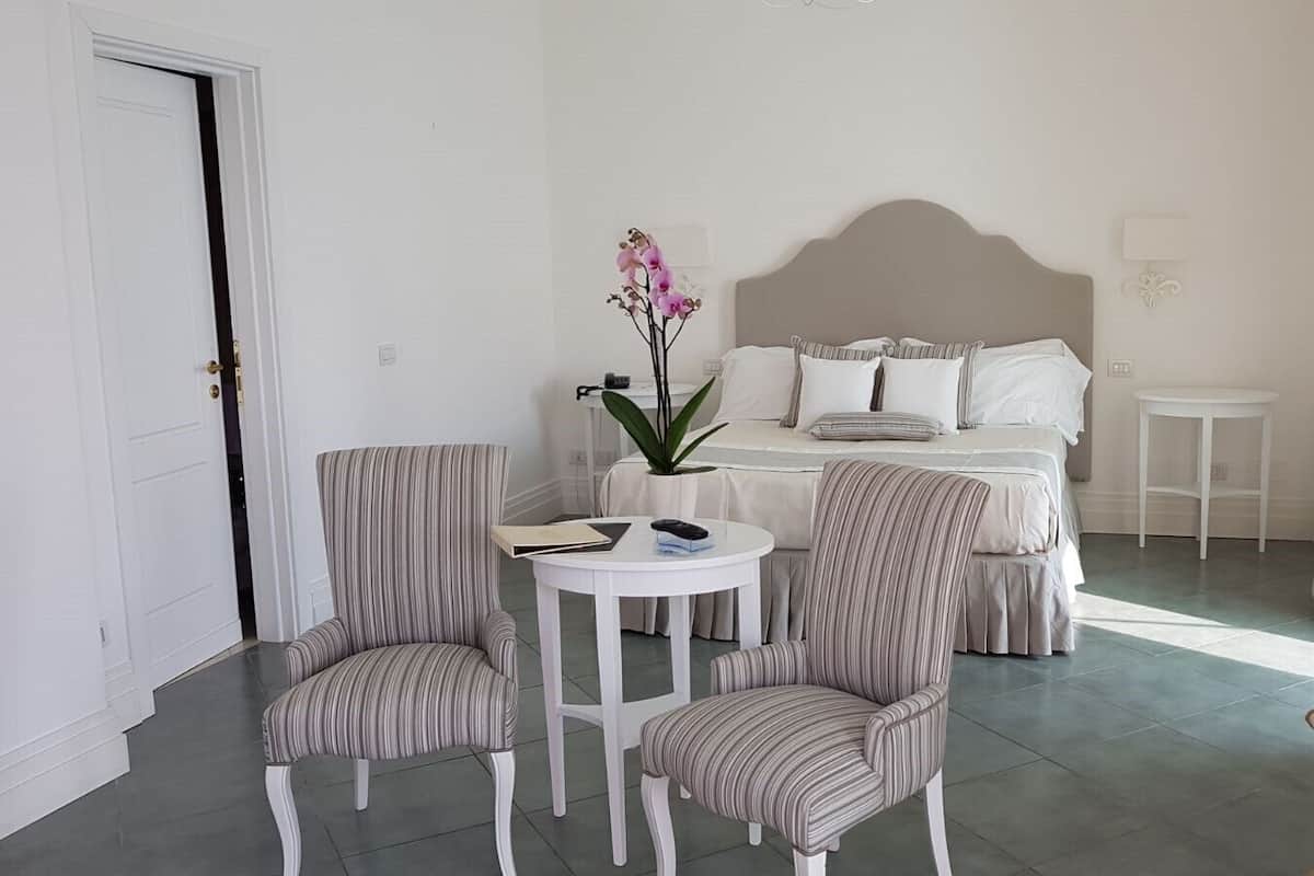 Quarto de hotel com uma cama, duas poltronas listradas, uma mesa redonda pequena com um livro e um controle remoto, uma orquídea em vaso e uma mesa de cabeceira com uma luminária.