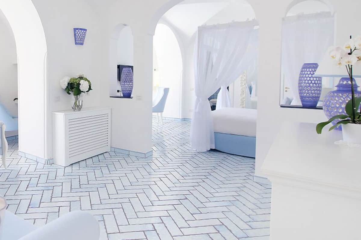 Um quarto claro e arejado com piso em padrão herringbone branco e azul, móveis brancos e vista para o mar através de uma varanda aberta.