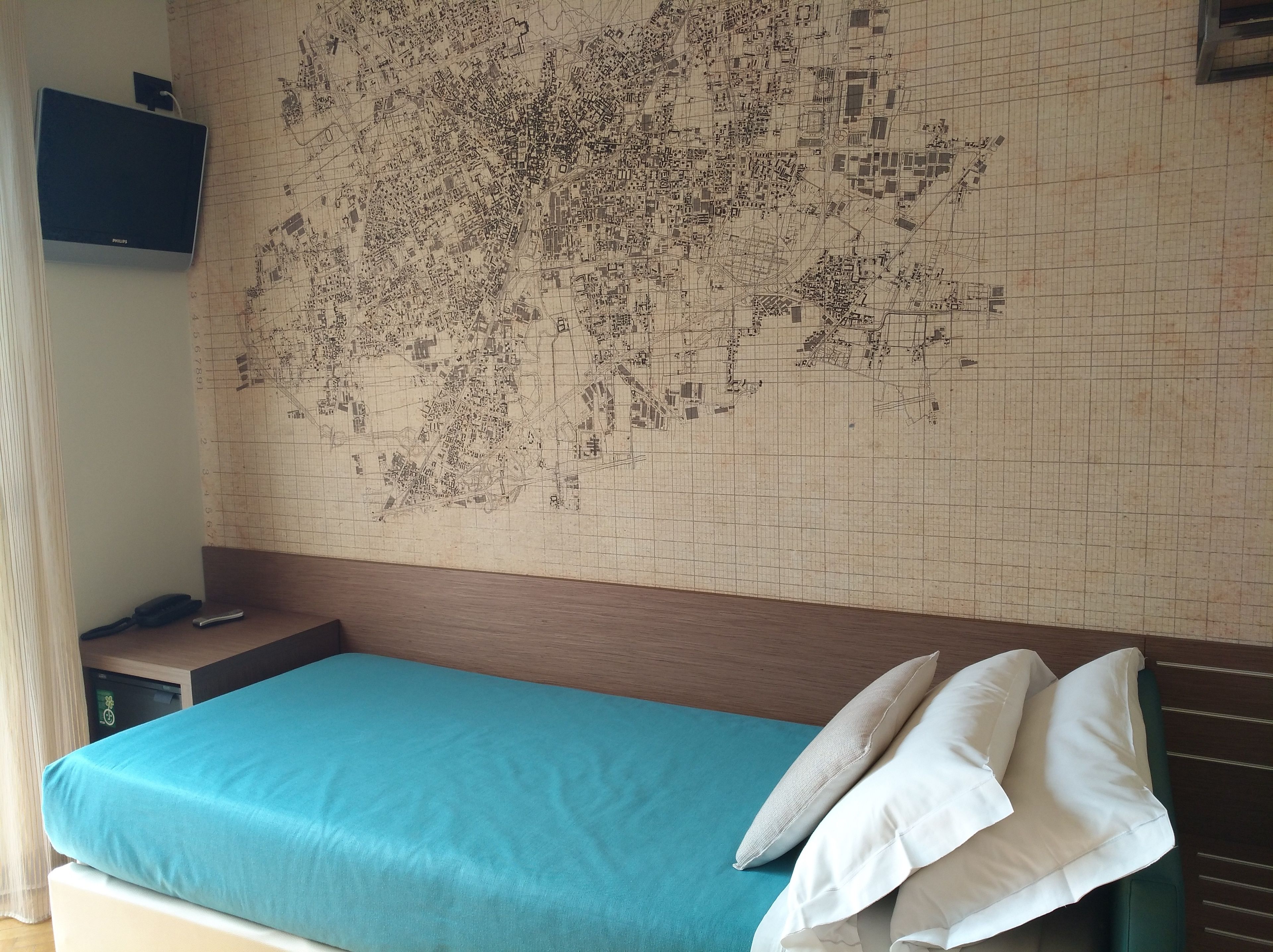 Um quarto com uma cama, um mapa na parede, uma mesinha e uma televisão presa à parede.