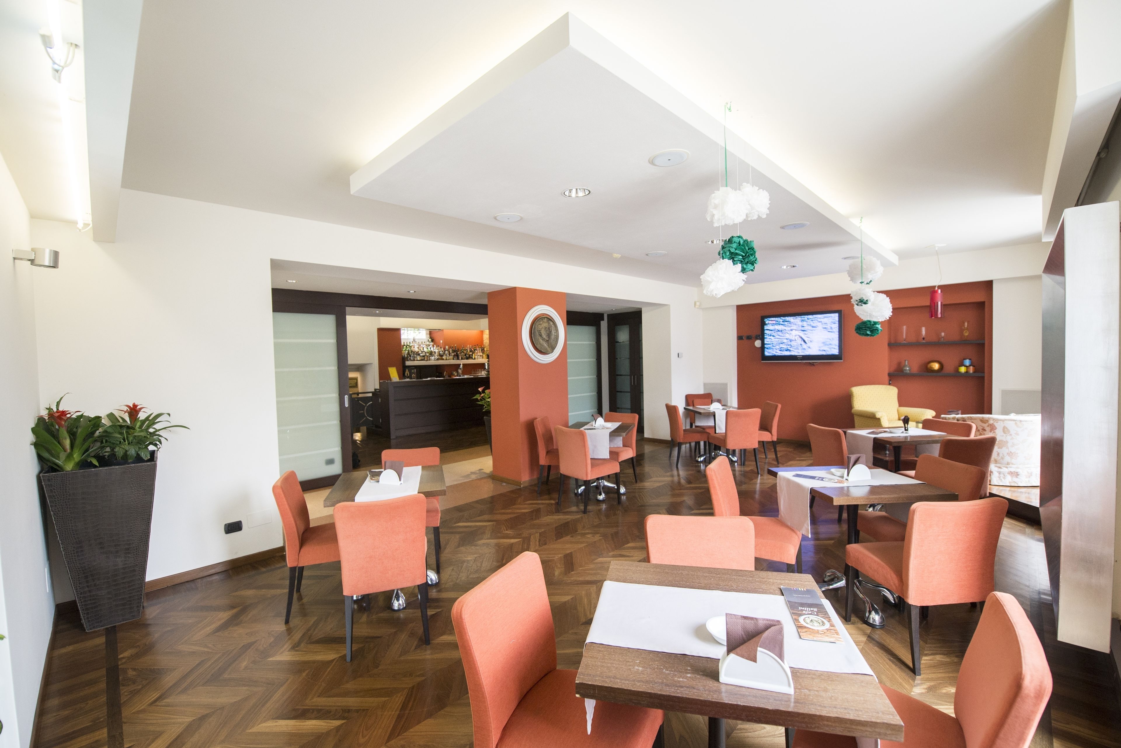 Uma área de refeições moderna com cadeiras e mesas laranja, um bar ao fundo e uma televisão fixada na parede.