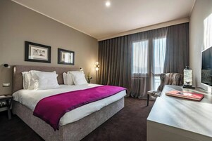Quarto de hotel com cama, escrivaninha, cadeira, televisão e cortinas.
