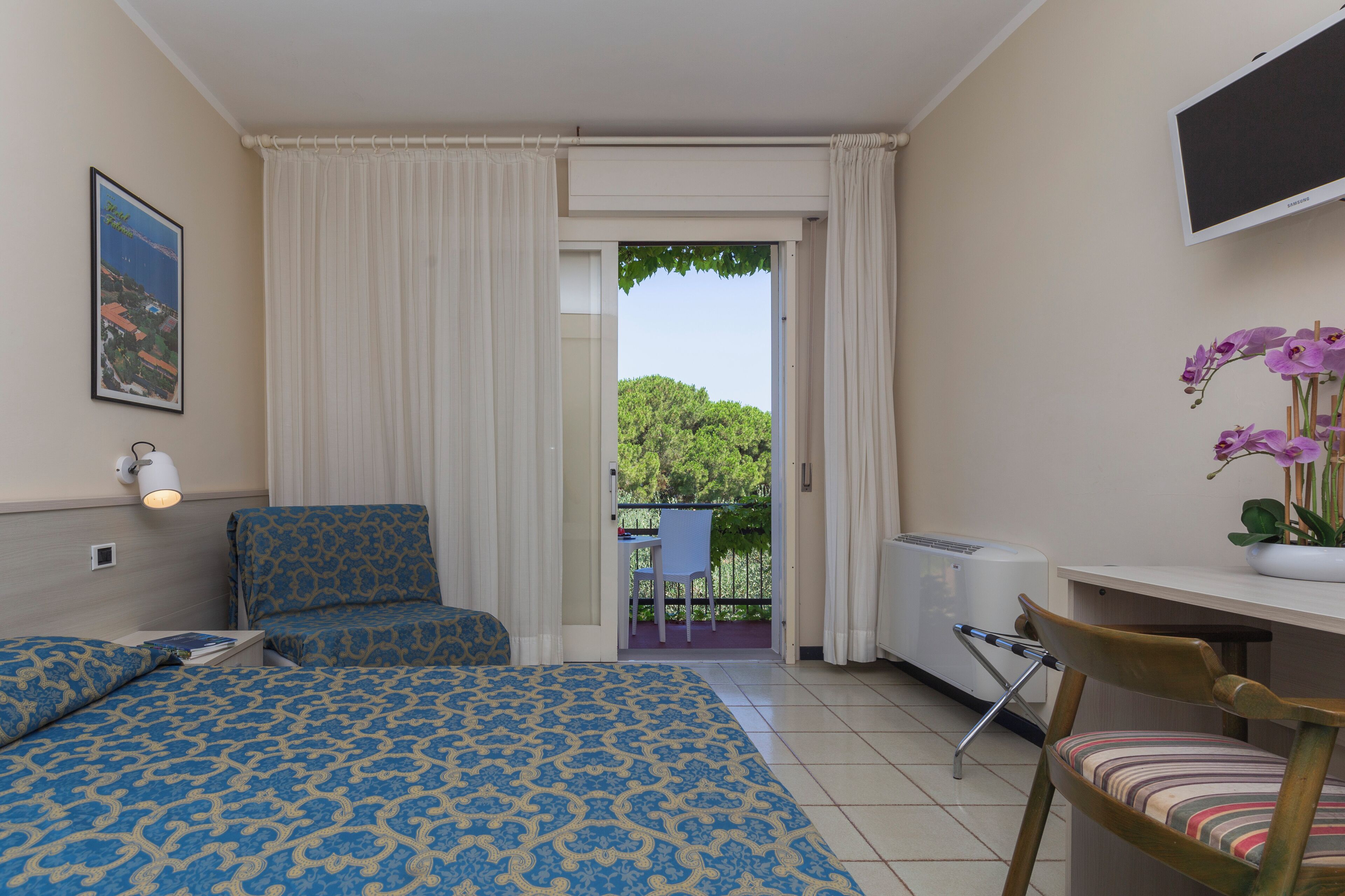 Quarto de hotel com cama, poltrona, escrivaninha, televisão e varanda com vista para o verde.