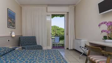 Quarto de hotel com cama, poltrona, escrivaninha, televisão e varanda com vista para o verde.