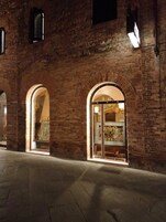 Um edifício de tijolos com arco e entrada por uma porta de vidro, iluminado por luzes externas.