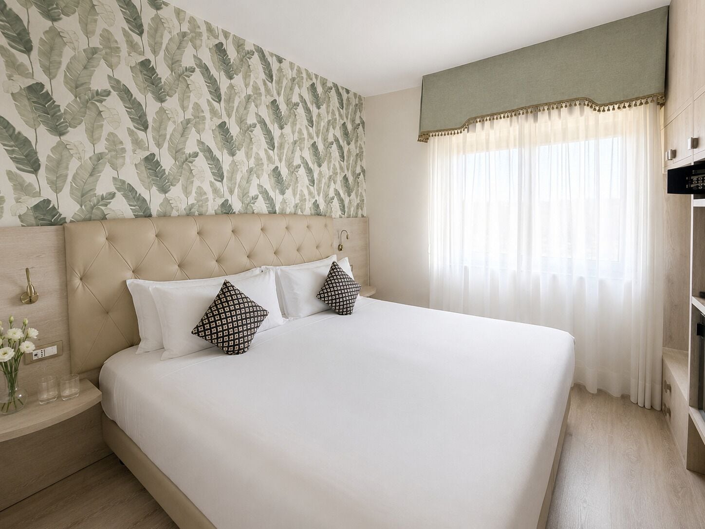 Quarto de hotel com uma cama grande, cabeceira, mesa de cabeceira com um vaso de flores e uma janela com cortinas.