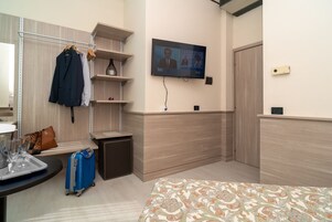 Quarto de hotel com uma cama, televisão na parede, guarda-roupa, secretária com um conjunto de copos e uma mala.