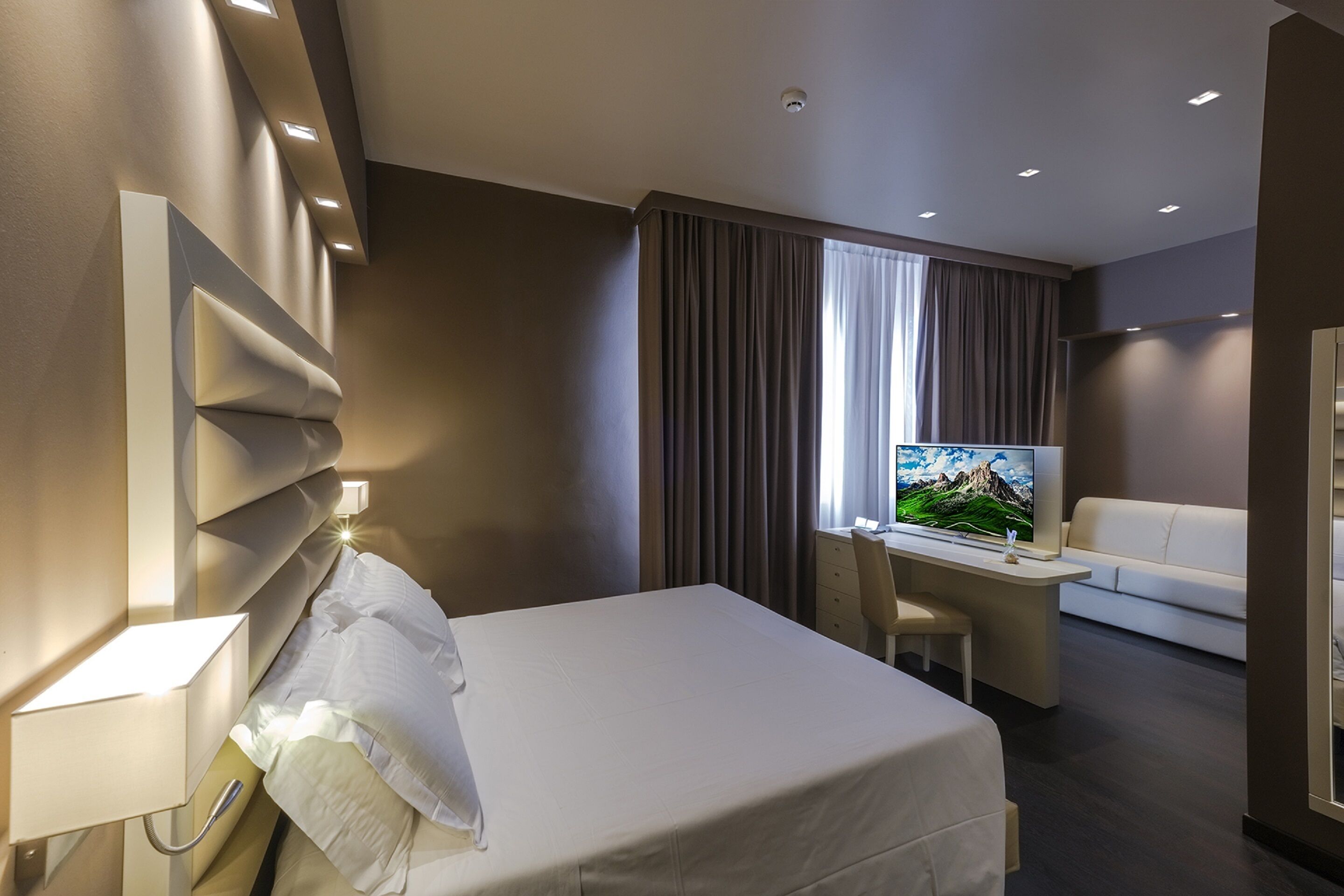 Quarto de hotel moderno com cama, escrivaninha com computador, televisão e sofá.